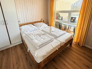 Quarto com cama de madeira, roupa de cama branca, janela com cortinas e vista para móveis de exterior.