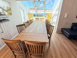 Uma sala de jantar com uma mesa de madeira comprida, cadeiras de madeira, um sofá preto e vista para um jardim através de uma parede de vidro.