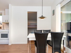 Uma sala de jantar com uma mesa branca e cadeiras pretas, uma luminária pendente e vista pela porta de vidro.