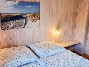 Quarto com duas camas, parede de madeira, quadro de uma praia e luminária fixada na parede.