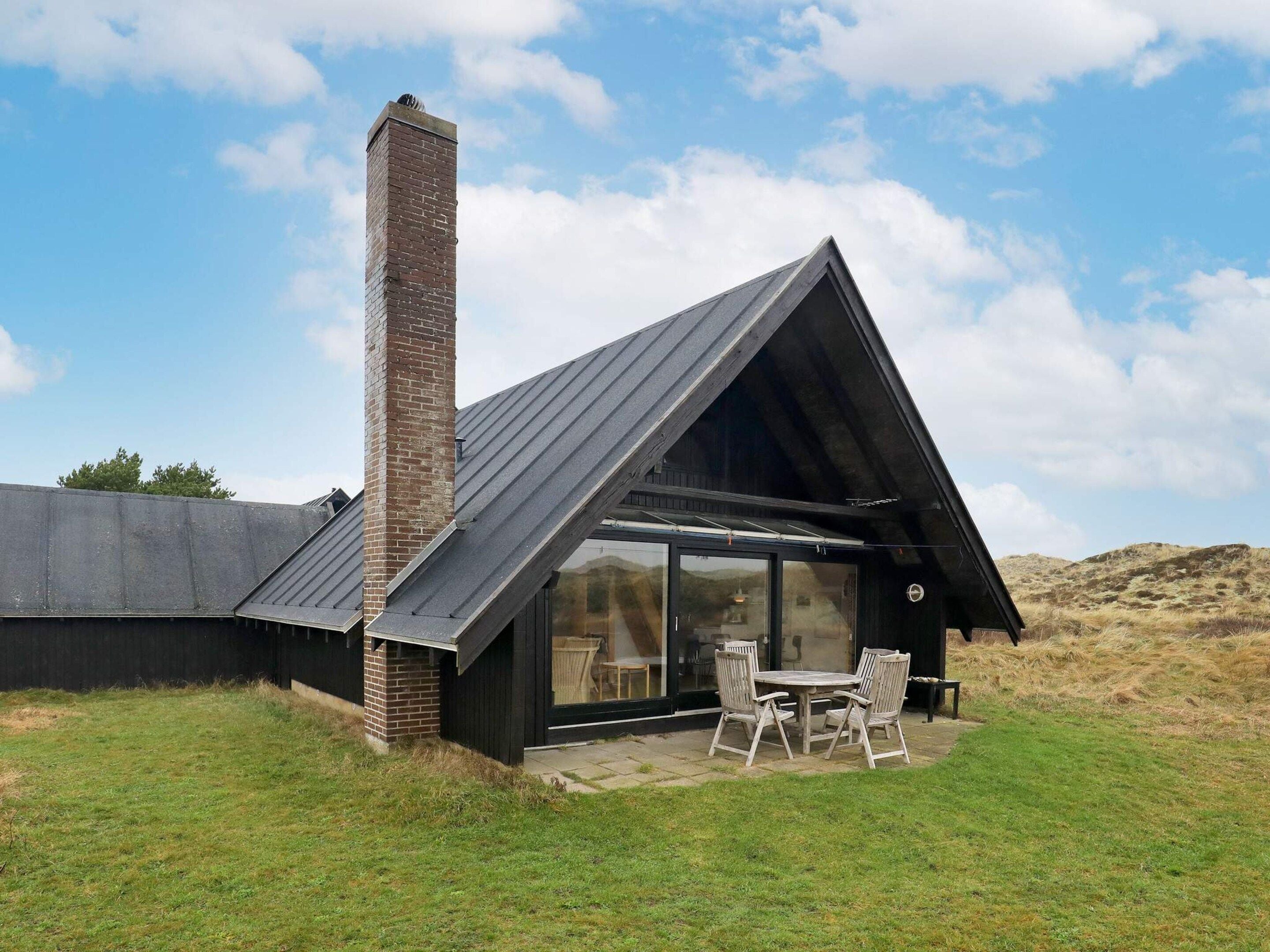 Uma casa moderna com telhado inclinado, chaminé de tijolos e porta de vidro que dá para uma área de estar externa com mesa e cadeiras.