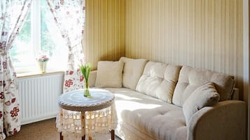 Quarto aconchegante com um sofá bege, uma mesa redonda pequena com toalha de renda e cortinas florais.