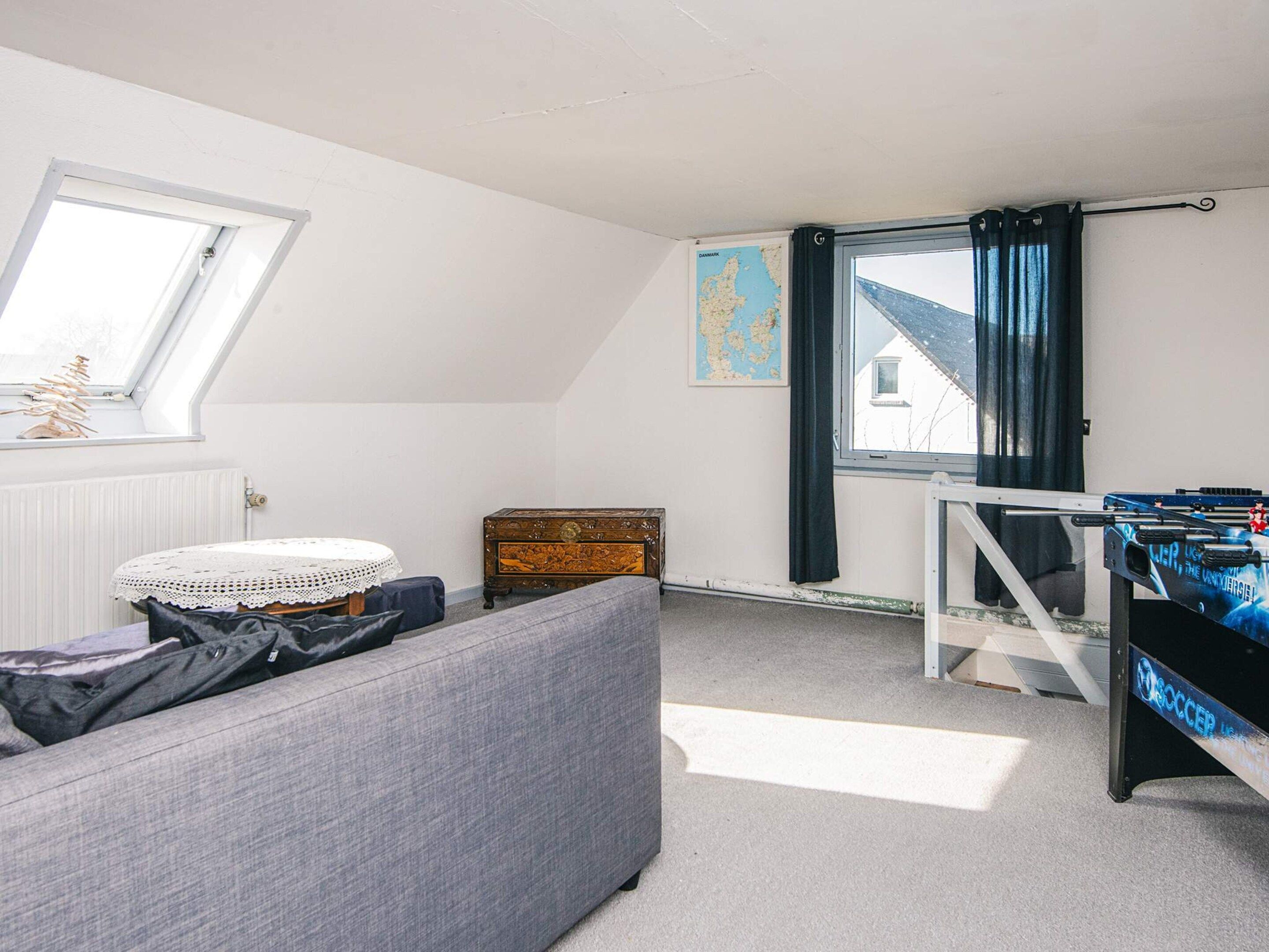 Um quarto com um sofá cinza, um pequeno baú de madeira, uma mesa com toalha rendada, uma janela com vista para uma casa e uma mesa de futebol.