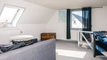 Um quarto com um sofá cinza, um pequeno baú de madeira, uma mesa com toalha rendada, uma janela com vista para uma casa e uma mesa de futebol.