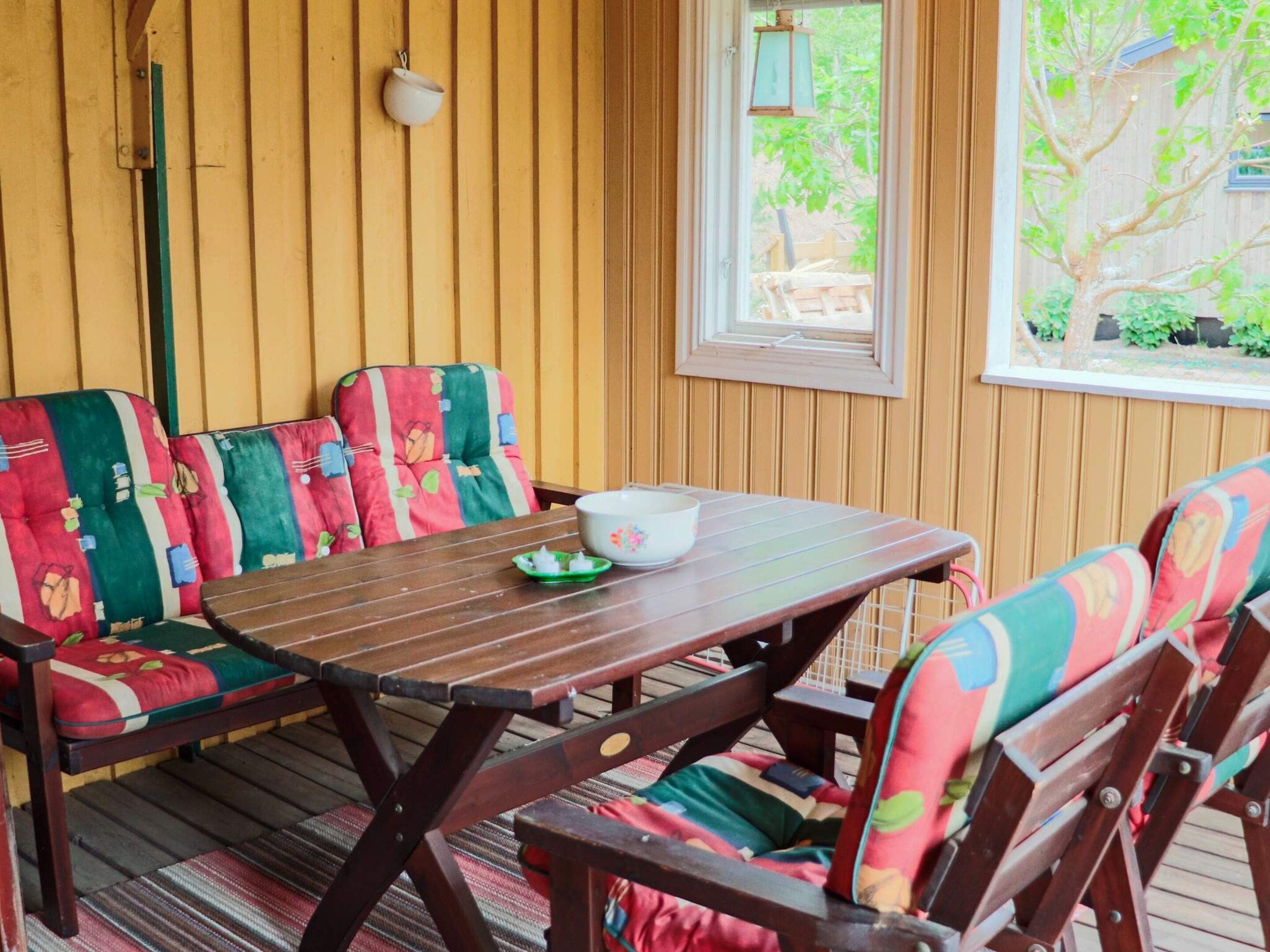 Um deck de madeira com mesa, cadeiras e um banco estofado, e uma janela com vista para o verde.