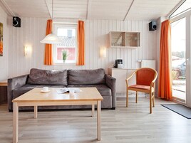 Uma sala de estar moderna com um sofá, uma mesa de madeira e uma cadeira. Há uma janela com cortinas, um alto-falante fixado na parede e um radiador.