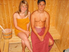 Duas pessoas sentadas em um vestiário de sauna de madeira, com um banco e um balde de madeira.