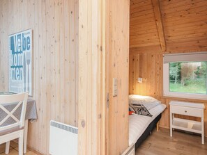 Interior de uma cabana de madeira com uma cama, uma mesa pequena, uma cadeira, um cartaz e uma janela com vista para árvores.