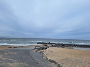 Uma praia com um molhe rochoso, um caminho pavimentado e uma faixa de areia.