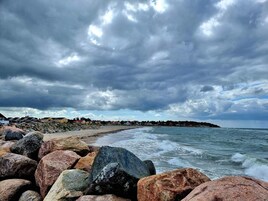 Uma linha de costa rochosa com ondas, céu nublado e uma linha de costa distante com edifícios.