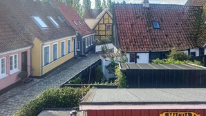 Uma vila europeia tradicional com ruas de calçada, edifícios históricos com telhados de terracota vermelha e um prédio vermelho em destaque com a placa 'MAGDA'.