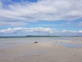Uma praia com areal, uma pequena rocha e uma faixa de terra distante sob um céu parcialmente nublado.