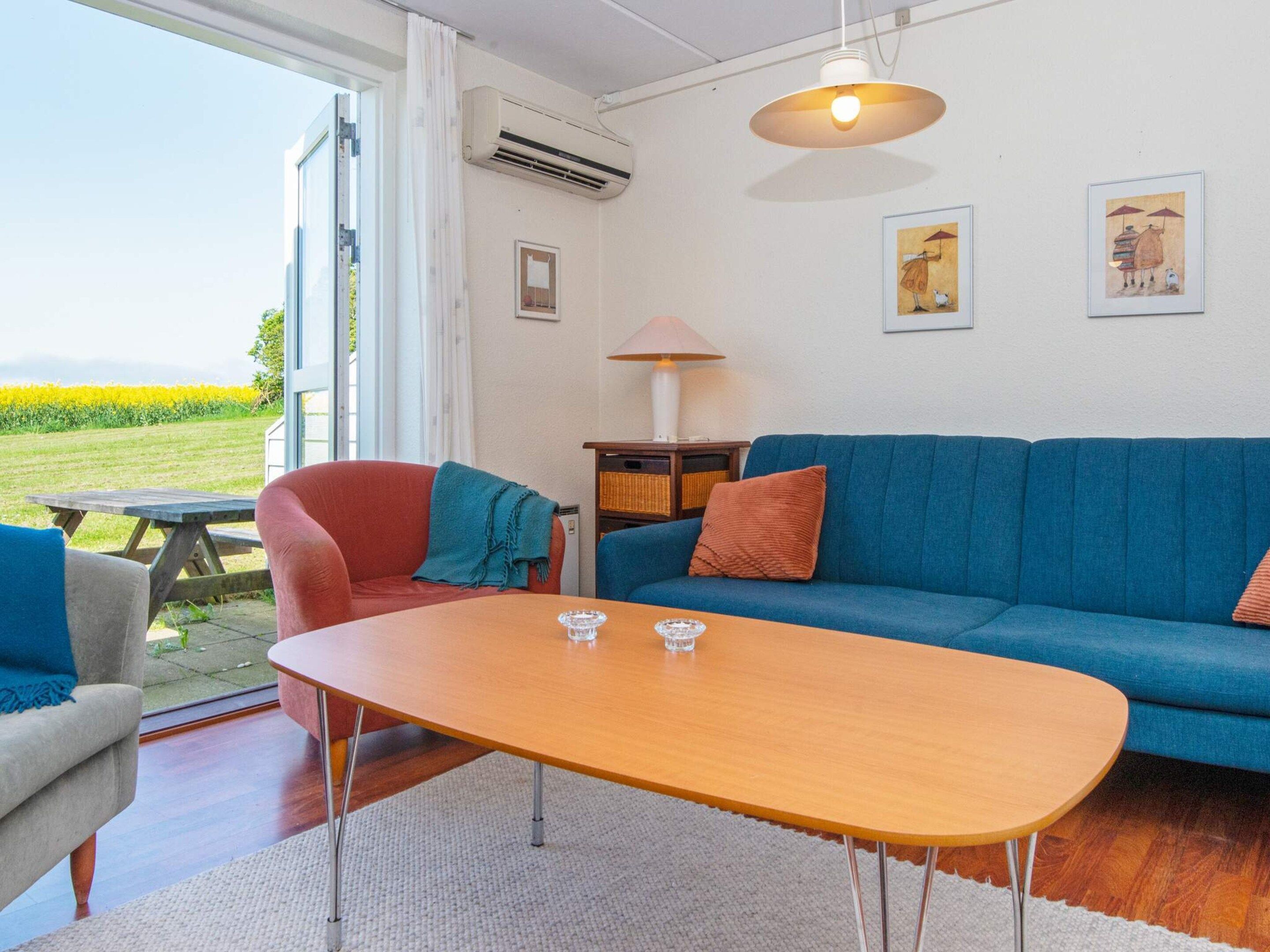 Sala de estar com sofá azul, poltrona vermelha, mesa de centro de madeira e vista para um campo verde pela janela.