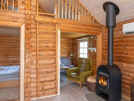 Interior de cabana de tronco com quarto no mezanino, uma poltrona verde, lareira e janela com vista.