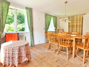 Uma sala de jantar com mesa e cadeiras de madeira, toalha de mesa estampada e janela com cortinas.
