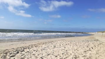 Uma praia arenosa com ondas, céu claro e um afloramento rochoso ao longe.