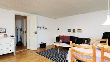 Uma sala de estar com piso de madeira, tapete preto e branco, sofá de couro preto, mesa branca e uma cadeira de madeira.