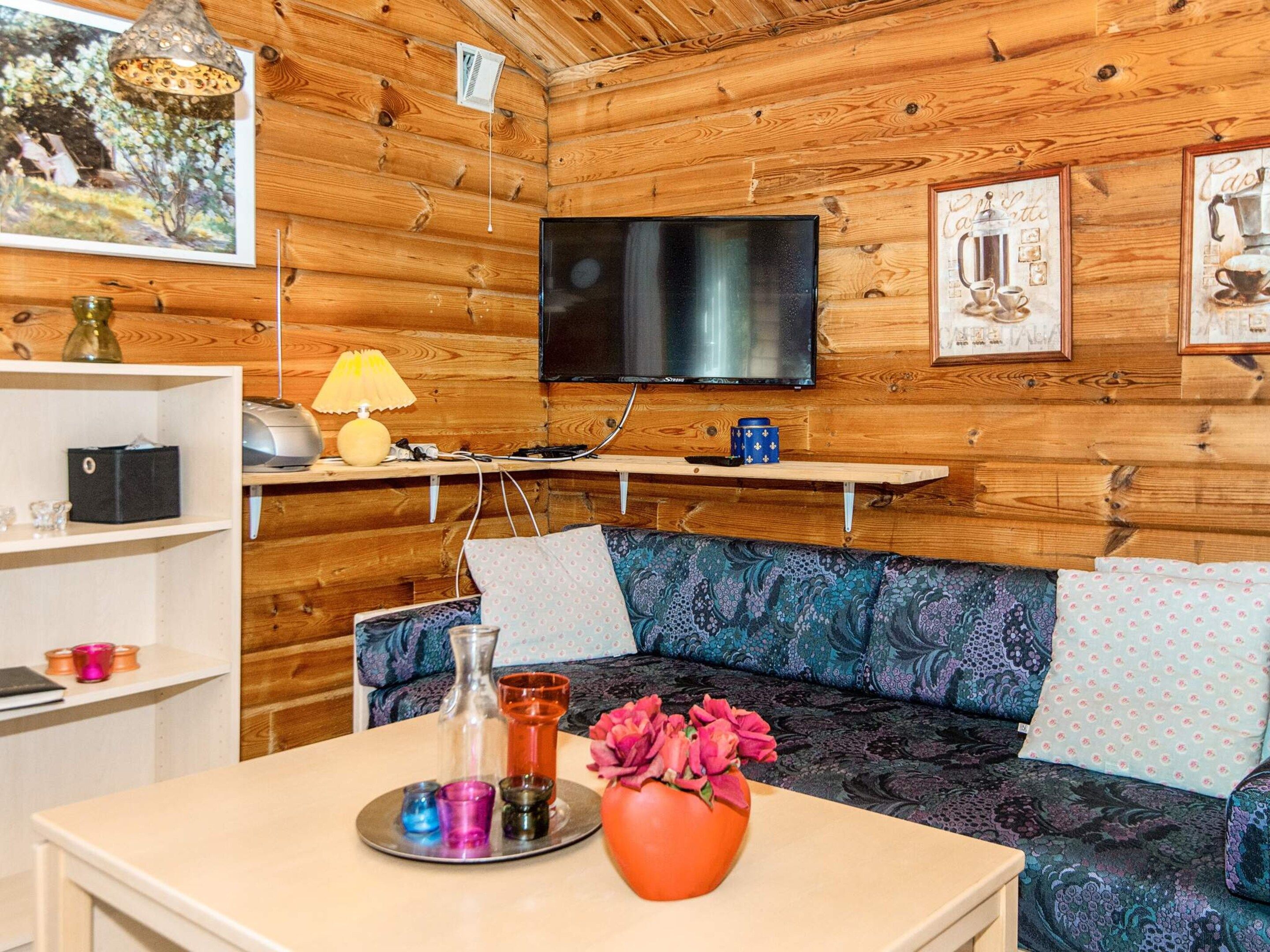 Interior acolhedor de uma cabana com parede de madeira, uma televisão sobre um sofá e uma mesa com um vaso de flores.