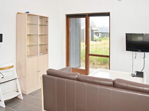 Quarto com sofá marrom, armário de madeira com prateleiras de vidro, televisão na parede e porta de vidro deslizante para área externa.