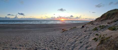 Uma praia com um pôr do sol, areia e uma duna gramada.