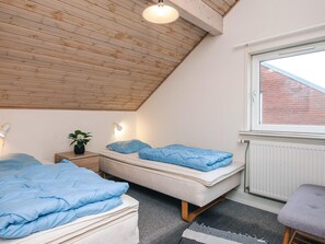 Quarto com teto inclinado, janela, radiador e mesa de cabeceira com abajur e planta.