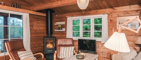Interior acolhedor de uma cabana com lareira, poltronas de couro, televisão e teto de madeira.