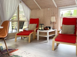 Quarto com duas cadeiras de madeira com almofadas vermelhas, uma mesa branca com uma luminária e vista pela janela.