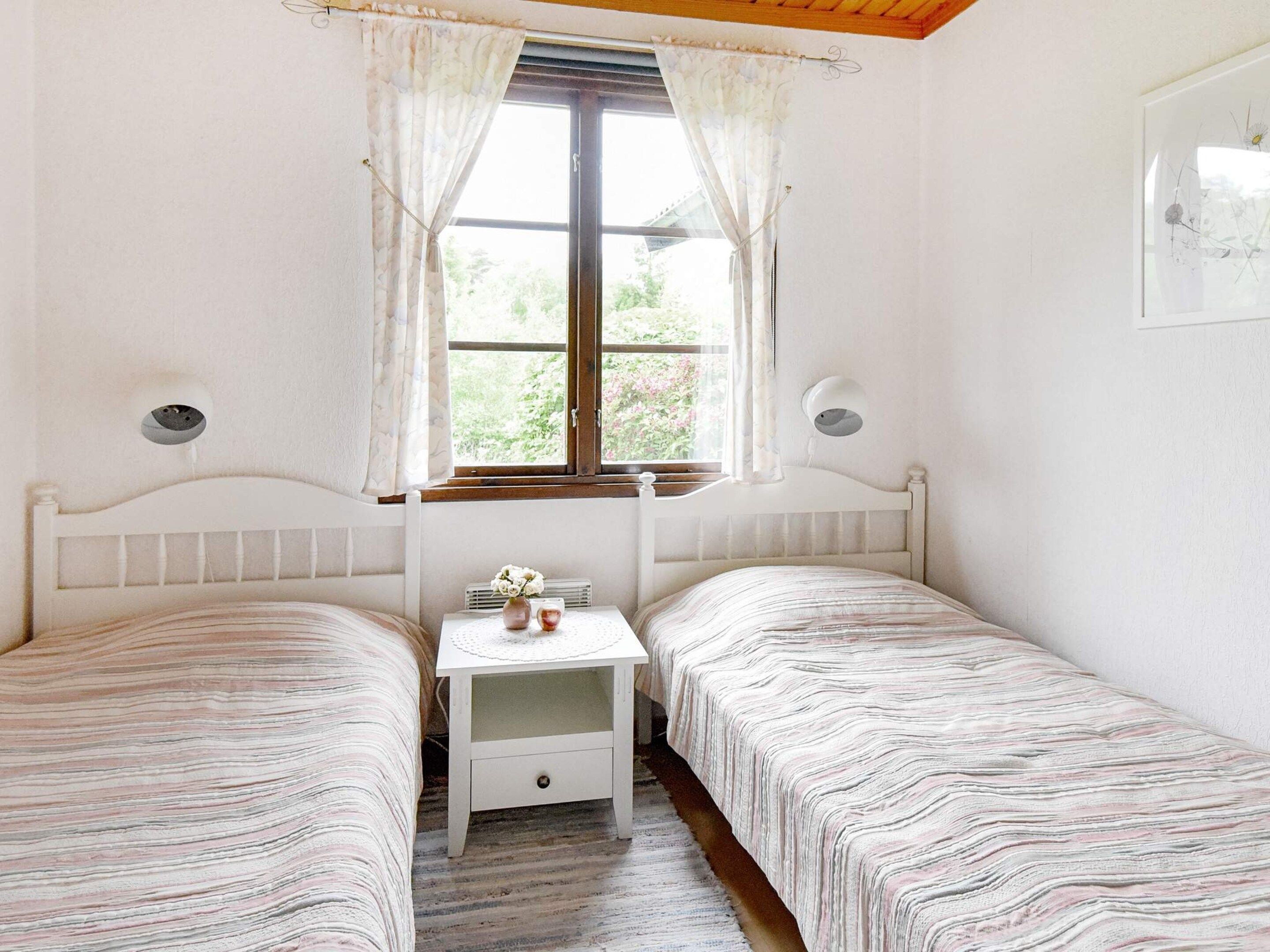 Quarto com duas camas de solteiro, uma pequena mesa de cabeceira com um vaso e abajur, e uma janela com cortinas.