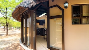 Uma casa com telhado de palha, cozinha visível por uma porta aberta, e um caminho de tijolos que leva à entrada.