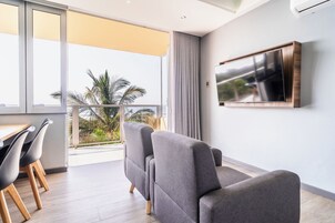 Uma sala de estar moderna com uma janela ampla, sofá cinza, mesa de jantar com cadeiras e uma televisão de tela plana na parede.