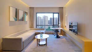 Quarto de hotel moderno com sofá, uma mesa redonda pequena, TV e uma grande janela com vista para a cidade.