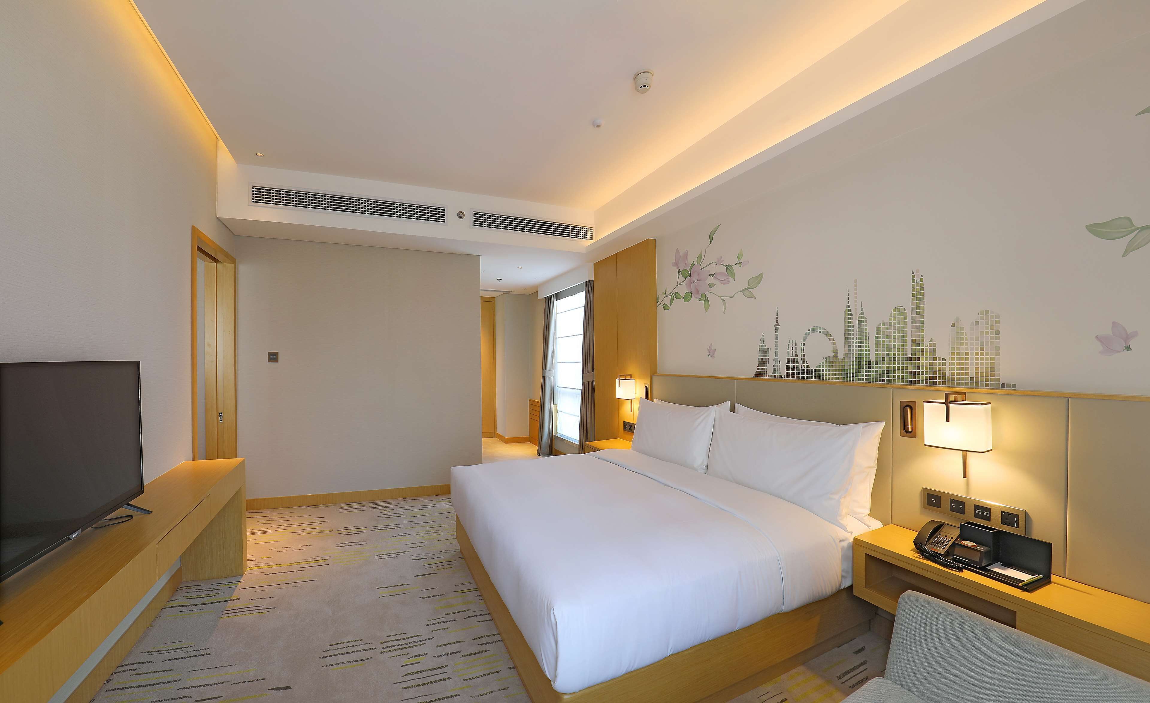 Quarto de hotel com uma cama, mesa de cabeceira com abajur, televisão e um mural com vista para a cidade.
