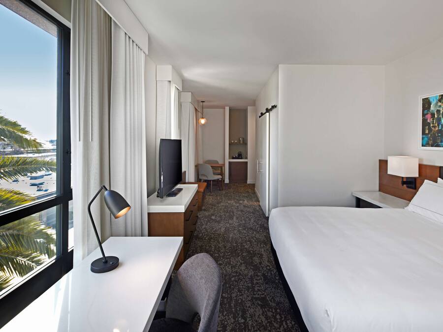 Quarto de hotel com uma cama grande, uma mesa com computador e luminária, uma cadeira, uma TV e vista para palmeiras pela janela.