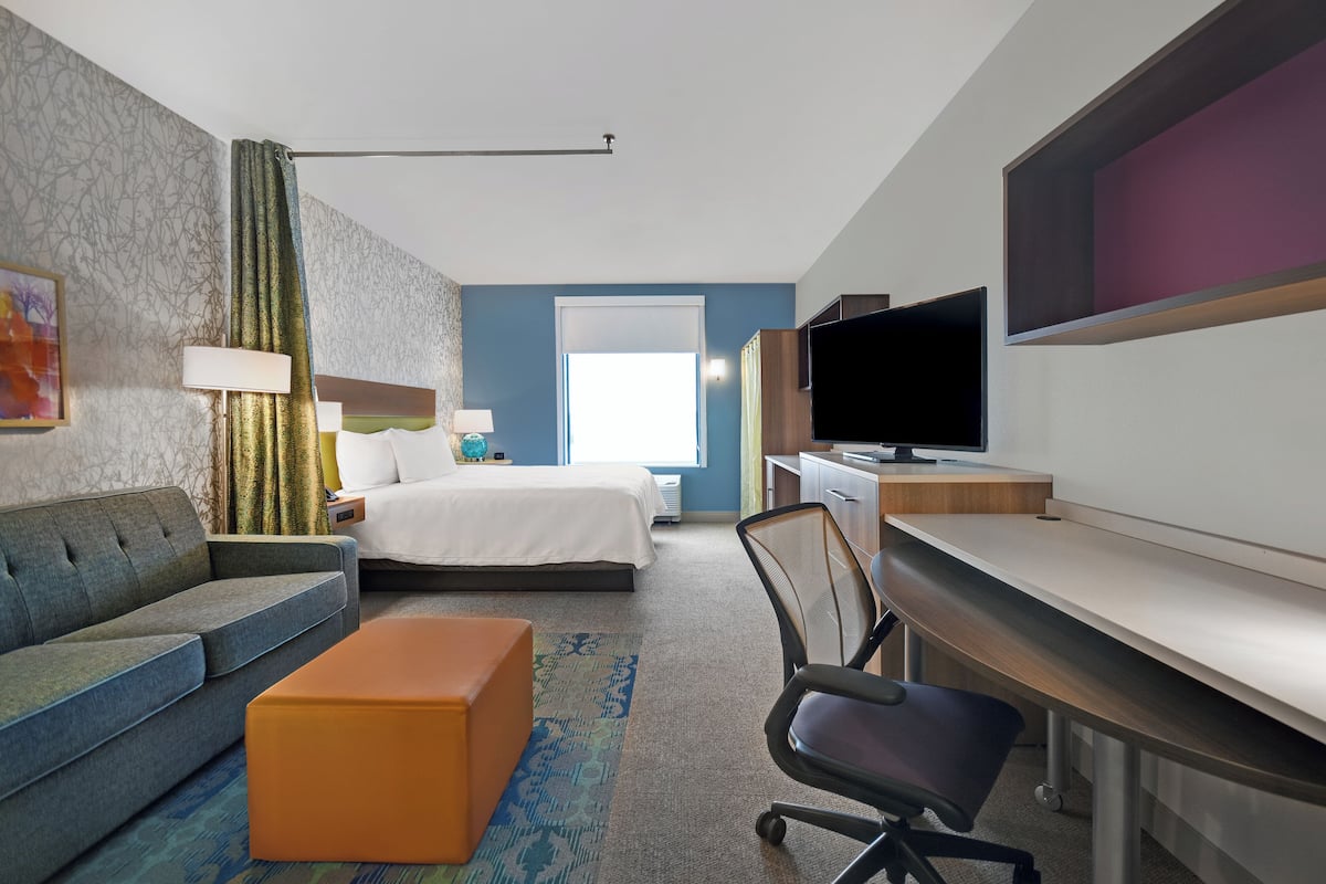 Quarto de hotel com cama, escrivaninha, cadeira, sofá, televisão e janela.