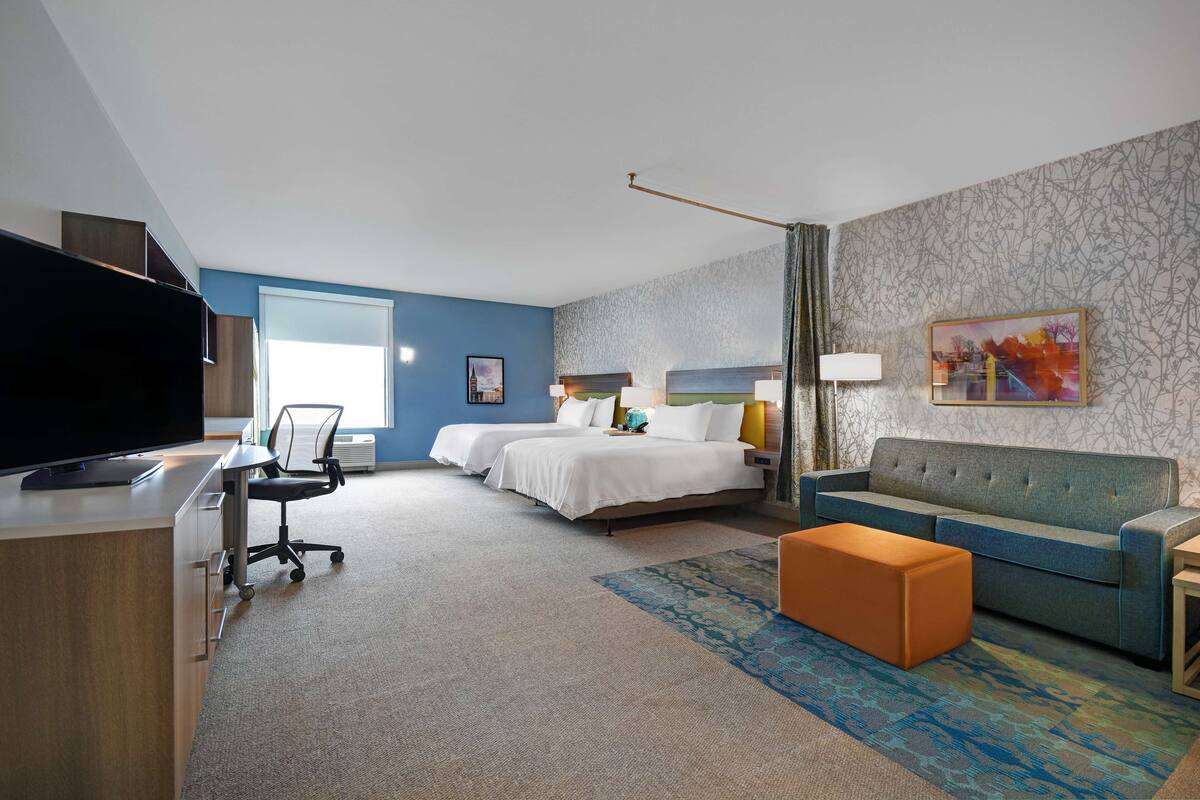Quarto de hotel com escrivaninha, duas camas, um sofá e uma televisão.