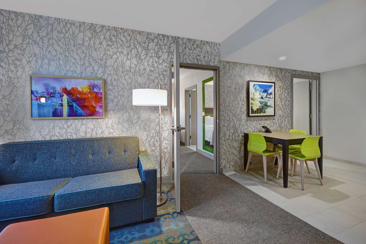 Quarto de hotel moderno com um sofá azul, uma mesa de jantar com cadeiras verdes e duas obras de arte emolduradas nas paredes.