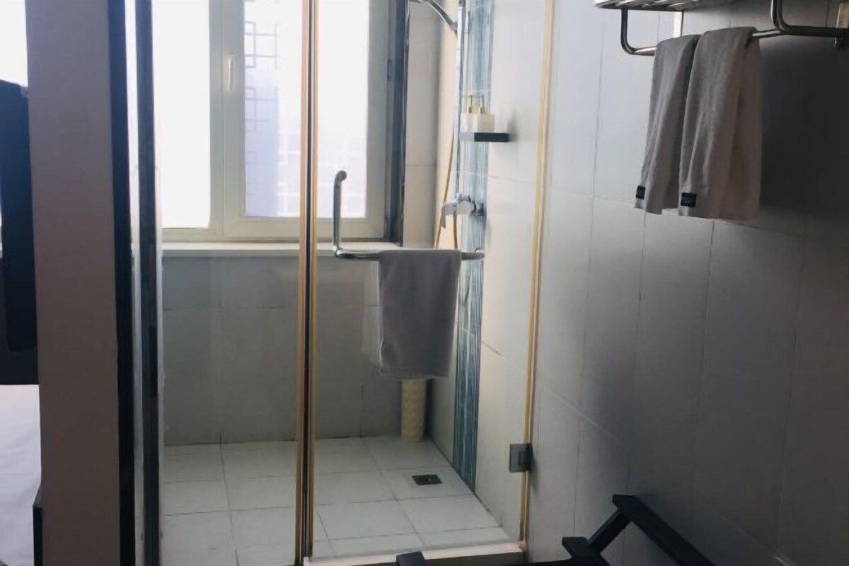 Pequeno box de chuveiro com porta de vidro, um porta-toalhas com toalha e uma janela com cortinas.