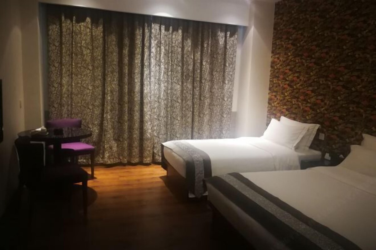 Quarto de hotel com duas camas, uma mesinha com uma cadeira roxa e uma janela grande com cortinas estampadas.