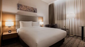 Quarto de hotel com uma cama grande, duas mesas de cabeceira com abajures, um criado-mudo, uma cadeira, uma pequena mesa com um vaso e uma janela grande com cortinas.