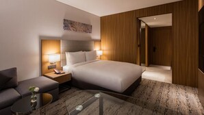 Quarto de hotel moderno com uma cama grande, abajures, um pequeno sofá e uma mesa de centro de vidro.