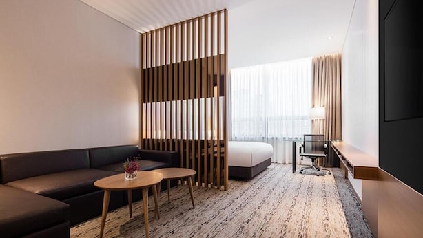 Quarto de hotel moderno com sofá modular, mesa de centro redonda, cama com cabeceira branca, escrivaninha com cadeira e uma televisão grande na parede.