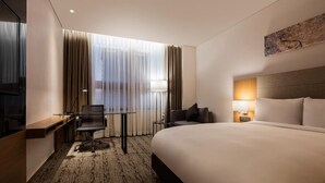 Quarto de hotel com uma cama grande, uma escrivaninha com cadeira, uma mesinha e uma luminária.