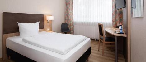 Quarto de hotel com cama, escrivaninha, cadeira, televisão e janela com cortinas.