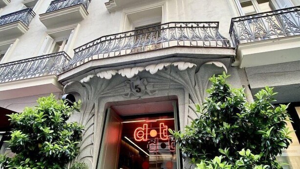 Entrada de hotel com letreiro de neon, plantas em vasos grandes e design arquitetônico clássico.