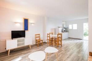 Uma sala de estar moderna com TV, cadeiras de madeira, uma mesa pequena e uma área de cozinha.