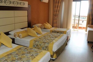 Quarto de hotel com três camas, cada uma com roupa de cama amarela e estampas florais. Há uma janela grande com cortinas, e é visível uma varanda pela janela.