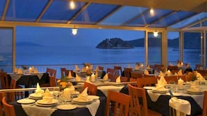 Restaurante com mesas preparadas para refeições, amplas janelas com vista para o mar e uma ilha ao longe, e design de interiores moderno.