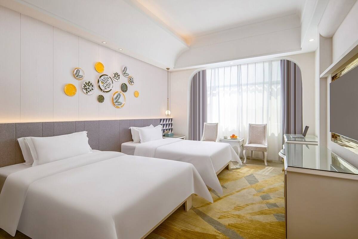 Quarto de hotel com duas camas, uma escrivaninha, uma cadeira e uma janela grande com cortinas.