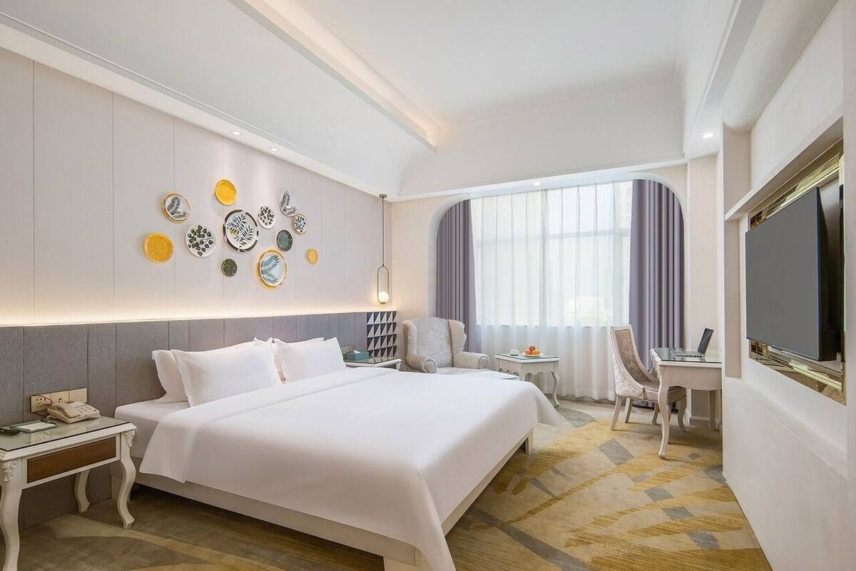 Quarto de hotel com uma cama grande, uma mesa de escritório com um laptop, uma televisão e uma área de estar com uma cadeira e uma mesa pequena.