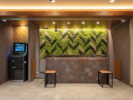 Uma área de recepção com um quiosque digital, dois bancos de madeira e uma parede com um padrão verde e preto.